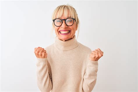 고립된 흰색 배경 위에 터틀넥 스웨터와 안경을 착용한 중년 여성은 팔을 들고 눈을 뜨고 성공을 축하하고 놀랐습니다 우승자 컨셉 매우 기쁜에 대한 스톡 사진 및 기타 이미지