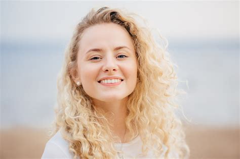 곱슬 머리와 행복한 젊은 귀여운 여성의 초상화를 닫고 카메라에 기꺼이 직접 보이는 넓은 미소를 가지고 해변 배경에 대한 포즈 사람 아름다움과 긍정적 인 감정 개념 개성