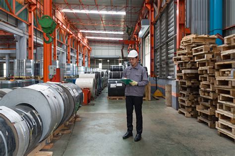 안전모와 보호장갑을 낀 작업자가 공장에 서서 문서 클립보드를 들고 자료를 확인하고 있습니다 환경은 금속 및 목재 팔레트 롤로 구성됩니다 20 29세에 대한 스톡 사진 및