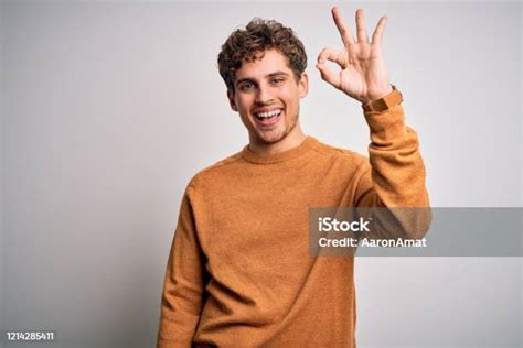젊은 금발 잘생긴 남자 곱슬 머리 흰색 배경 에 캐주얼 스웨터를 입고 긍정적 인 하고 확인 기호를 하고 손과 손가락 성공적인 식입니다 겨울에 대한 스톡 사진 및 기타 이미지