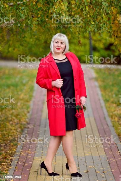 검은 드레스를 입은 예쁜 금발과 손에 장미가 달린 빨간 코트가 공원의 길에 서 있다 여성의 크기 플러스 Xxl 일반 계획 수직 뷰 가을에 대한 스톡 사진 및 기타 이미지