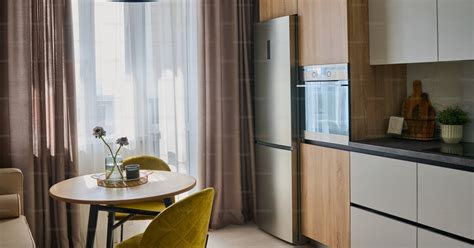 둥근 주방 테이블과 파스텔 커튼과 냉장고가 있는 창문 사이에 서 있는 겨자 노란색의 편안한 의자 2개 사진 Unsplash의 국산 부엌 이미지