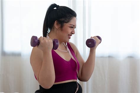 집에서 아령과 함께 운동을하는 젊은 과체중 여성 집에서 앉아있는 동안 손에 아령을 들고 목에 측정 테이프와 과체중 뚱뚱한 여자