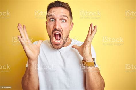 노란 고립 된 배경 위에 캐주얼 한 흰색 티셔츠를 입은 젊은 잘생긴 남자는 미친 축하와 팔을 들고 눈을 뜨고 흥분 비명을 지르며 성공을 위해 놀랐습니다 우승자 컨셉 T