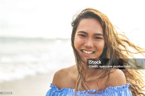 긴 갈색 물결 모양의 머리를 가진 젊은 밀레니얼 필리핀 여성의 초상화는 파도와 파도와 파도가 서핑 보드를 들고 물 근처 바다의 가장자리에 모래에 서 캘리포니아에서 물 해변을