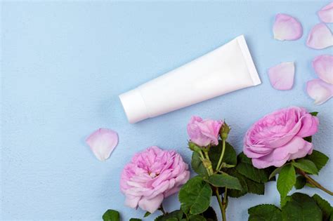 파란색 배경에 얼굴 손 바디 크림이있는 흰색 플라스틱 튜브와 핑크 다마스크 장미 새싹 모형 상단 보기 자연미용 제품