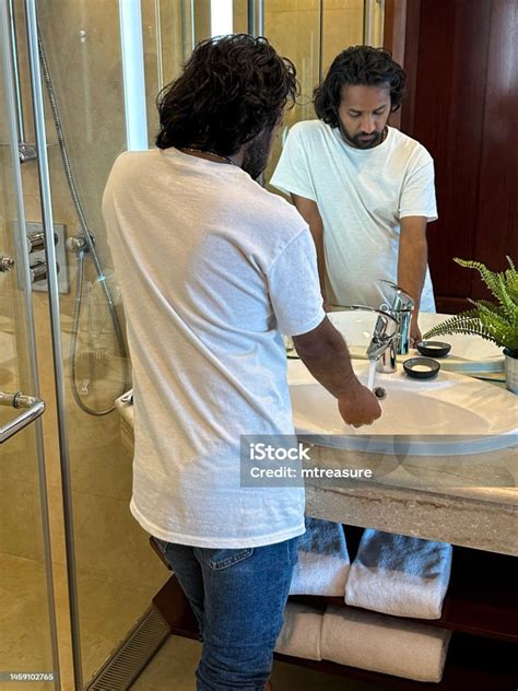 인도 남자의 거울에 비친 반사의 클로즈업 이미지는 흐르는 물에서 손을 씻는 고급 욕실의 싱크대에 서 있었고 대리석 욕실 카운터의 금속 냄비에 인공 양치류 식물 흐르는 수도