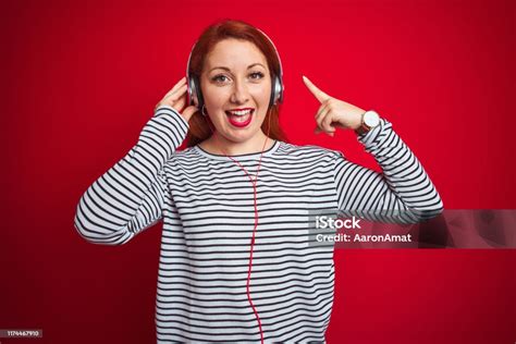 젊은 빨간 머리 여자 듣고 음악을 사용 하 여 헤드폰 을 통해 빨간 고립 된 배경 매우 행복 가리키는 손과 손가락 가리키기에 대한 스톡 사진 및 기타 이미지 Istock