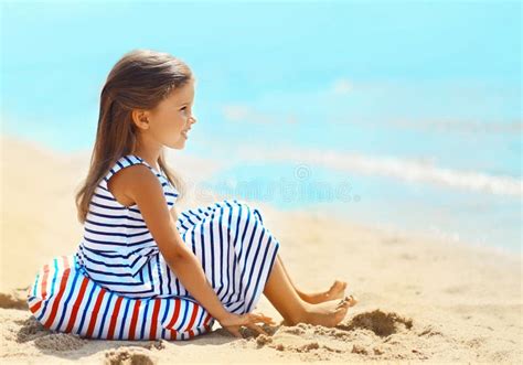 Маленькая девочка сидя на пляже песка около моря в лете Стоковое Фото изображение