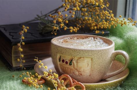 아침 식사로 향기로운 아침 커피 베이지 색 세라믹 컵에 큰 거품창과 니트 모헤어에 서 있다 인근에는 독서책과 노란 미모사 꽃이 있습니다 0명에 대한 스톡 사진 및 기타