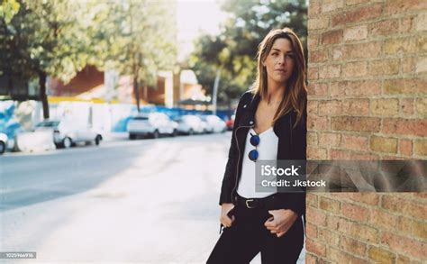 가죽 자 켓에 벽돌 벽에 기대어 거리에 서 있는 동안 제쳐두고 찾고 입고 긴 머리를 가진 젊은 세련 된 학생 여자의 길이 초상화 잠겨있는 매력적인 여자 옆으로 찾고 가을에