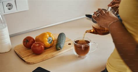 찻주전자를 들고 뜨거운 음료를 머그잔에 붓는 젊은 여성의 클로즈업 스마트폰과 야채가 있는 주방 카운터 옆에 서 있다 사진 Unsplash의 컵 이미지