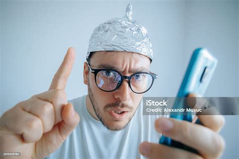알루미늄 호일 모자를 쓴 남자가 방에서 스마트폰을 들고 불신을 표현하고 있다 35 39세에 대한 스톡 사진 및 기타 이미지 35 39세 Brand Name Smart