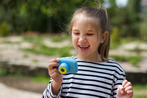 밝은 화창한 날에 공원에서 줄무늬 티셔츠를 입은 긴 금발 머리를 가진 웃는 소녀가 작은 카메라를 들고 사진을 찍고 그것을 들여다 봅니다 선택적 초점 개념에 대한 스톡 사진
