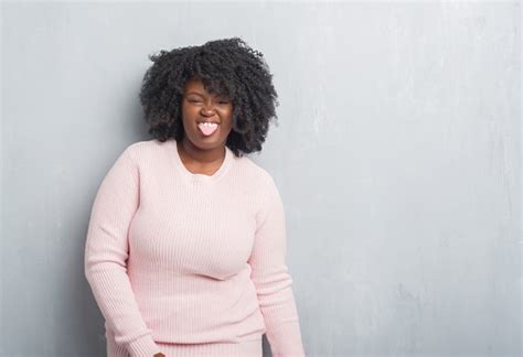 젊은 아프리카계 미국인 플러스 크기 여자 혀를 내민 겨울 스웨터를 입고 회색 그런 지 벽에 재미 있는 표현을 함께 행복을 밖으로 감정 개념입니다 가을에 대한 스톡 사진 및