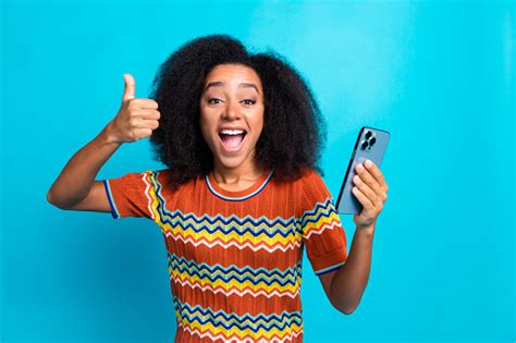 갈색 줄무늬 티셔츠를 입고 새로운 아이폰 15를 사용하여 파란색 배경에 고립 된 속도 엄지 손가락을 즐기는 젊은 재미 소녀 물결 모양의 헤어 스타일 사진 25 29세에 대한