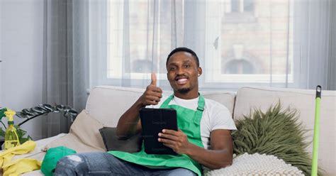 제복을 입은 아프리카계 미국인 남자가 소파에 앉아 디지털 태블릿을 손에 들고 카메라에 엄지손가락을 치켜세운 채 웃고 있다 남성 청소부는 현대적인 아파트에서 청소 과정에서
