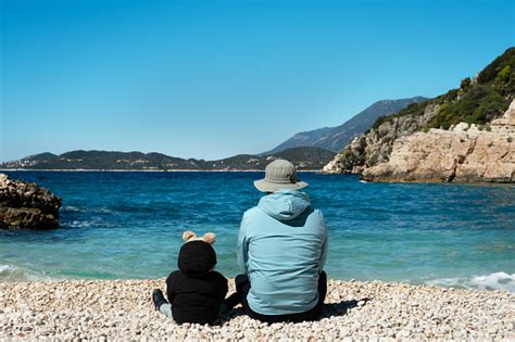 추운 날씨에 바다를 바라보며 해변에 앉아 따뜻한 옷을 입은 남자와 아기 아버지와 아이는 야외에서 자연 아름다운 장소에서 함께 시간을 보내고 뒤에서 보기 2명에 대한 스톡