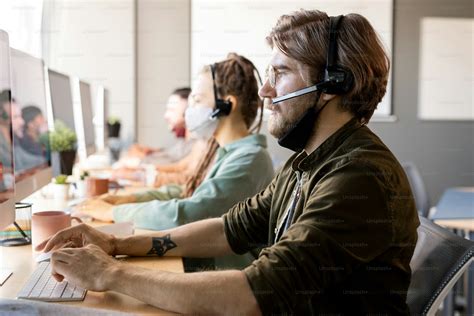 턱 아래에 보호 마스크를 쓴 젊은 수염 난 남성 오퍼레이터가 헤드셋을 착용한 동료들 앞에 앉아 고객을 상담하고 있다 사진 Unsplash의 컴퓨터 이미지