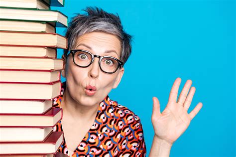 안경을 쓴 쾌활한 노인 여성이 열정적으로 손을 흔들며 생생한 파란색 배경을 배경으로 키 큰 책 더미 옆에 서 있습니다 50 54세에 대한 스톡 사진 및 기타 이미지 Istock