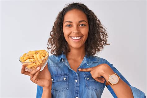 자신에게 손가락을 가리키는 깜짝 얼굴과 고립 된 흰색 배경을 통해 마카로니 파스타와 그릇을 들고 젊은 브라질 여자 가리키기에