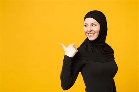 웃는 젊은 아라비아 무슬림 여자 히 잡 검은 옷 노란 벽 배경 스튜디오 초상화에 고립 된 손 손가락으로 가리키는 복사 공간을