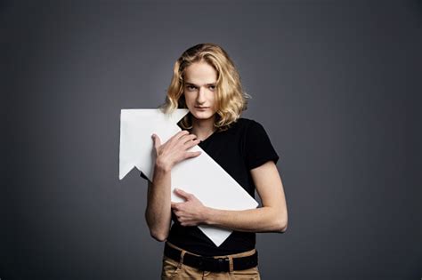 긴 금발 머리와 미소 얼굴을 가진 잘 생긴 젊은 남자가 회색 배경에 오른쪽을 가리키는 흰색 화살표를 포옹 20 24세에 대한 스톡 사진 및 기타 이미지 Istock