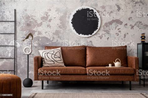 현대적인 갈색 소파 모의 포스터 프레임 말린 꽃이 있는 꽃병 Pouf 및 세련된 개인 액세서리가 있는 거실 인테리어의 현대적인 디자인 회색 콘크리트 벽입니다 가정 장식 템