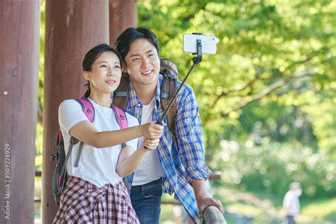 한국의 옛날 전통 가옥의 처마와 녹색 숲을 배경으로 여행하며 스마트폰으로 다양한 포즈를 셀카로 찍는 젊은 아시아 한국 남녀 모델 Stock 사진 Adobe Stock