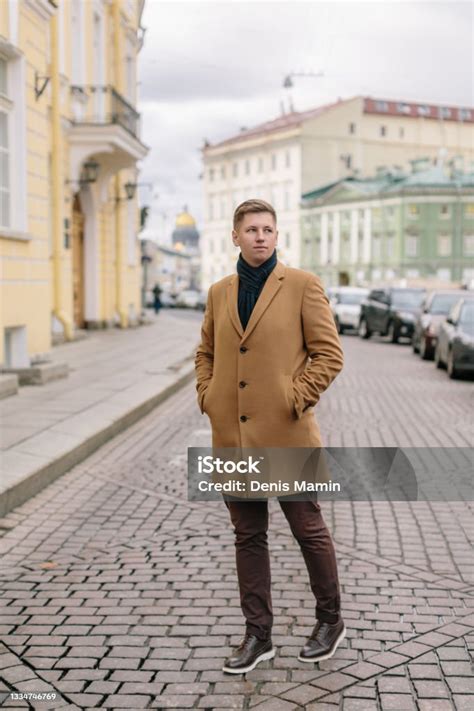 베이지 색 코트를 입은 매력적인 금발 백인 남성은 목에 짙은 파란색 스카프를 묶고 갈색 바지와 어두운 가죽 신발을 주머니에 넣습니다 25 29세에 대한 스톡 사진 및 기타