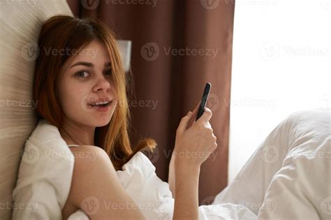 Woman With Mobile Phone Lies In Bed And Window Blanket Curtains