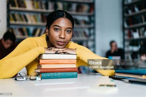 코스 작업을 작성하고 숙제를하기위한 많은 문학 교과서와 바탕 화면에 도서관에 앉아있는 동안 카메라를보고 아름다운 아프리카 계 미국인 학생의 절반 길이 초상화 아프리카계 미국