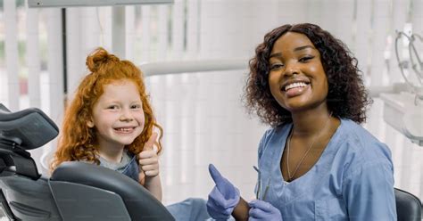 카메라를 보고 있는 쾌활한 미소를 짓는 아프리카 여성 치과의사와 치과 의자에 앉아 엄지손가락을 치켜든 작은 빨간 머리 소녀 환자의 전면 모습 치아 건강 구강 관리의 개념