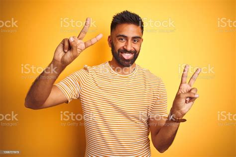 고립된 노란색 배경 위에 서있는 티셔츠를 입은 젊은 인도 남자가 승리의 표지판을 하고 있는 양손의 손가락을 드러내며 혀로