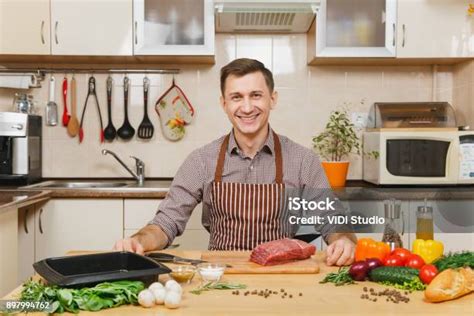 잘생긴 백인 젊은이 야채와 함께 테이블에 앉아 앞치마에 나무 표면 멋진 주방의 전체 빛 부엌에 돼지고기 쇠고기 또는 양고기 고기 스테이크를 준비 하는 가정에서 요리 가구에