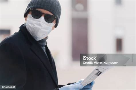 코로나 바이러스 건강 관리 보호 및 검역 개념 진지한 남자는 모자 선글라스 일회용 의료 마스크 및 고무 장갑을 착용하고 어딘가에 집중하고 신문을 읽고 야외에서 포즈를