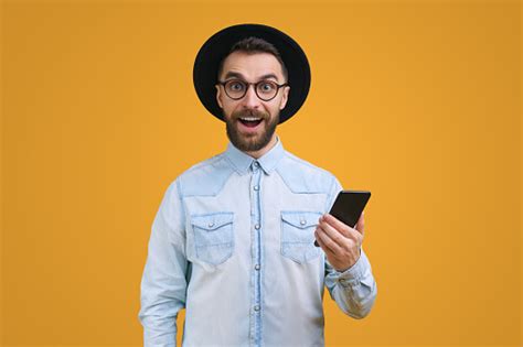 트렌디한 안경과 스마트폰을 손에 들고 모자를 쓰고 포즈를 취하는 흥분한 얼굴 표정으로 잘생긴 수염을 가진 남자의 스튜디오 초상화 Championship에 대한 스톡 사진 및