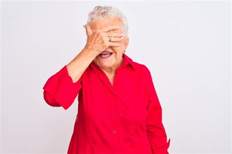 고립 된 흰색 배경 위에 서있는 빨간 캐주얼 셔츠를 입은 선임 회색 머리 여성은 놀라움을 위해 눈을 가리는 얼굴에 손으로 웃고 있습니다 블라인드 개념 가리기에 대한 스톡