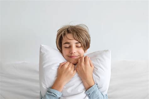 하얀 베개를 껴안고 눈을 감고 하얀 벽 배경에 앉아 있는 잘생긴 웃는 십대 소년 십대 건강한 생활 방식 수면 루틴 휴식 좋은 수면 시간 개념 Mental Wellbeing에