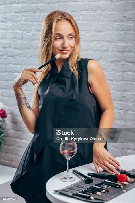 검은 옷을 입은 젊은 성인 여성이 팔을 벌리고 메이크업 브러쉬와 레드 와인 한 잔을 들고 있다 그녀는 프레임과 포즈를 진지하게 바라본다 20 29세에 대한 스톡 사진 및