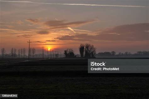 겨울철에 부분적으로 안개가 자욱한 시골 환경과 수평선에서 볼 수있는 구름 다양한 나무의 가시적 인 실루엣 오렌지색으로 칠해진 하늘 위에 해질녘 0명에 대한 스톡 사진 및