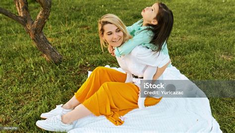 공원에서 그녀의 엄마를 포용 행복 한 아이의 측면 보기 귀여운 딸 편승 그녀의 젊은 어머니는 녹색 잔디 야외에서 흰색 담요에 앉아 어머니의 날입니다 모성 및 유년기 개념