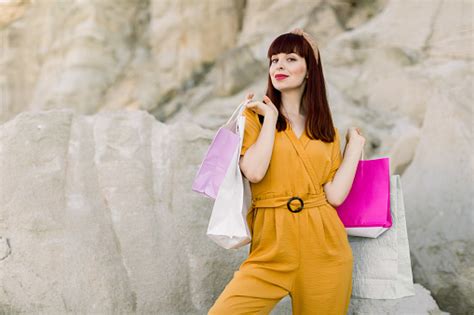 노란 여름 바지를 입고 어두운 직선 머리를 가진 아름다운 패션 소녀 그녀의 손에 화려한 쇼핑 백으로 서 사막 절벽이나 모래 채석장에서 포즈 가방에 대한 스톡 사진 및 기타