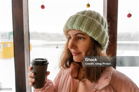 검은 종이 커피 컵을 들고 부드럽게 웃고 카페에 혼자 서서 손에 커피를 마시고 기분이 좋은 기분을 가진 것을 기쁘게 생각합니다 창 뒤의 겨울 갈색 머리에 대한 스톡 사진 및