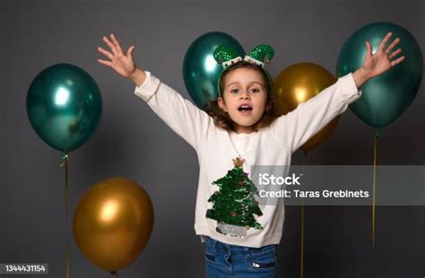 흰색 크리스마스 스웨터와 엘프 후프를 입고 아름다운 어린 소녀는 복사 공간으로 회색 배경위에 고립 된 다채로운 황금색과 녹색 아름다운 풍선에 맞서 팔을 들어 올리고 있습니다