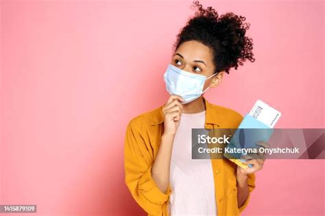 얼굴에 의료용 마스크를 쓴 펜시브 아프리카 계 미국인 곱슬 머리 소녀 여권과 여행 티켓을 들고 고립 된 분홍색 배경에 서서 옆을 바라보고 휴가를 꿈꾸며 20 24세에 대한