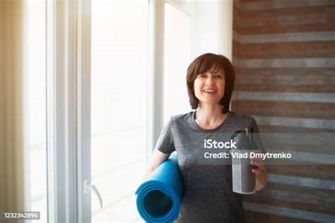 성인 적합 슬림 여자는 집에서 운동을 가지고있다 카메라와 미소를 바라보는 긍정적 인 쾌활한 시니어 여성 운동 후 단백질 쉐이크와 요가 매트를 손에 들고 있습니다 휴식을