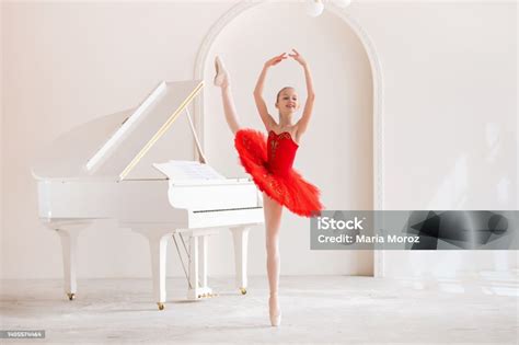 귀여운 어린 소녀는 프로 발레리나가되기를 꿈꿉니다 피아노 옆의 하얀 방에서 밝은 빨간색 투투를 입은 소녀가 뾰족한 신발을 신고 춤을 추고 있습니다 직업 학교 학생 12 13