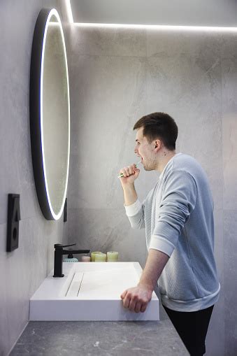 옅은 스웨터를 입은 남자가 현대식 욕실 세면대에 서서 동그란 거울에 비친 자신의 모습에 집중하며 양치질을 하고 있다 Led 조명에 대한 스톡 사진 및 기타 이미지 Istock