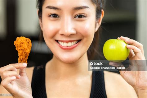 사과와 프라이드 치킨 정크 푸드를 먹는 운동복을 입은 아름다운 여성은 고칼로리 음식과 건강 위험 건강에 해로운 음식을 가진 배고픈 젊은 아시아 여성 가정 생활에 대한 스톡